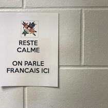 Une affiche blanche sur un mur blanc où il y est écrit : « Reste calme, on parle français ici ».